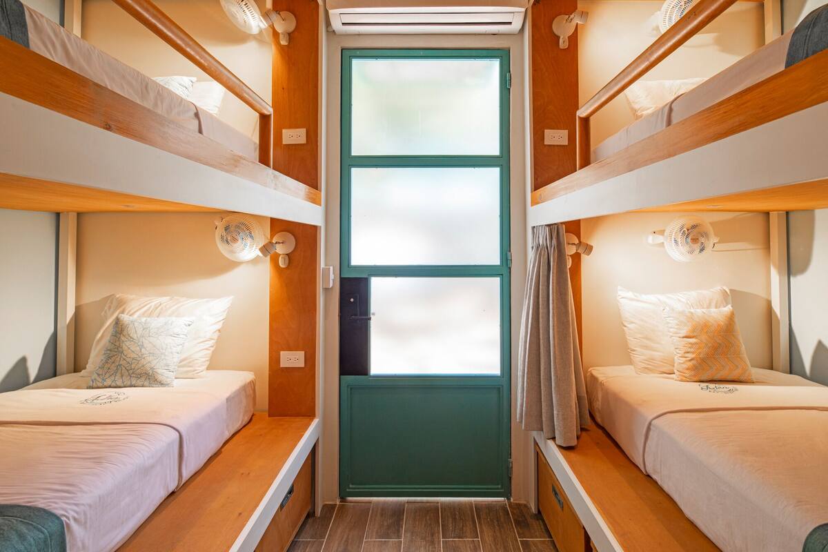 Um corredor estreito com beliches em ambos os lados, uma porta verde e ventiladores fixados na parede.