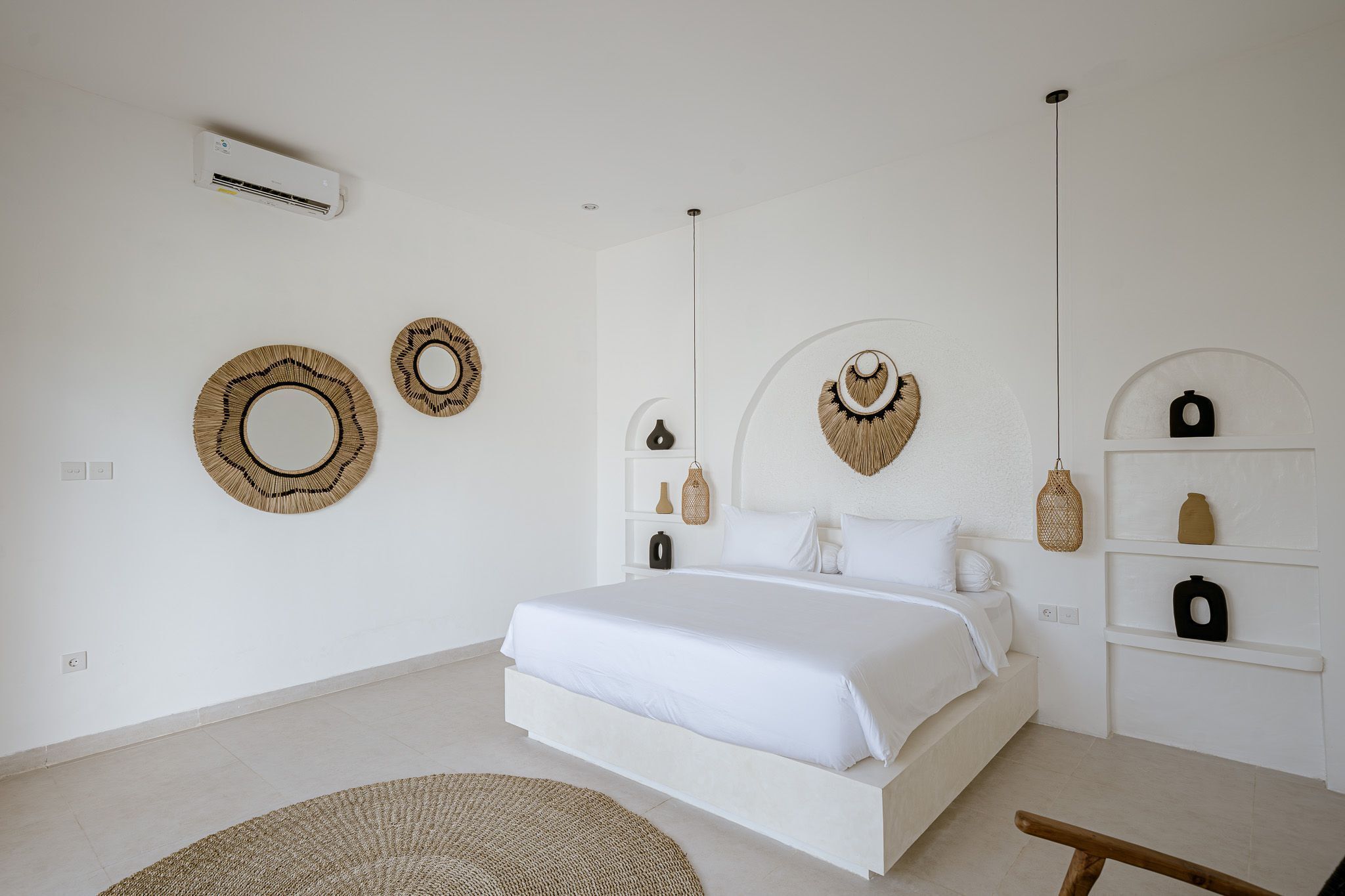 Quarto minimalista com uma cama grande, um espelho redondo e uma estante com objetos decorativos.