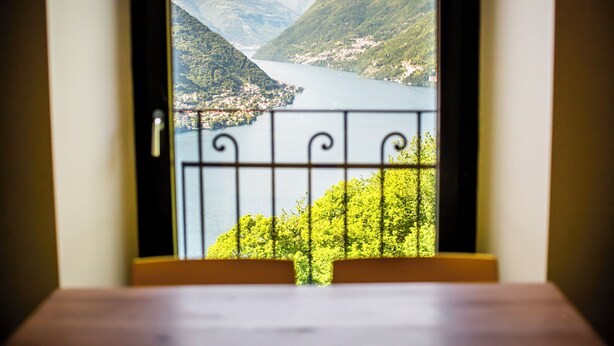 Vista de uma varanda com grade de metal, com vista para um lago rodeado por colinas e um pequeno povoado.
