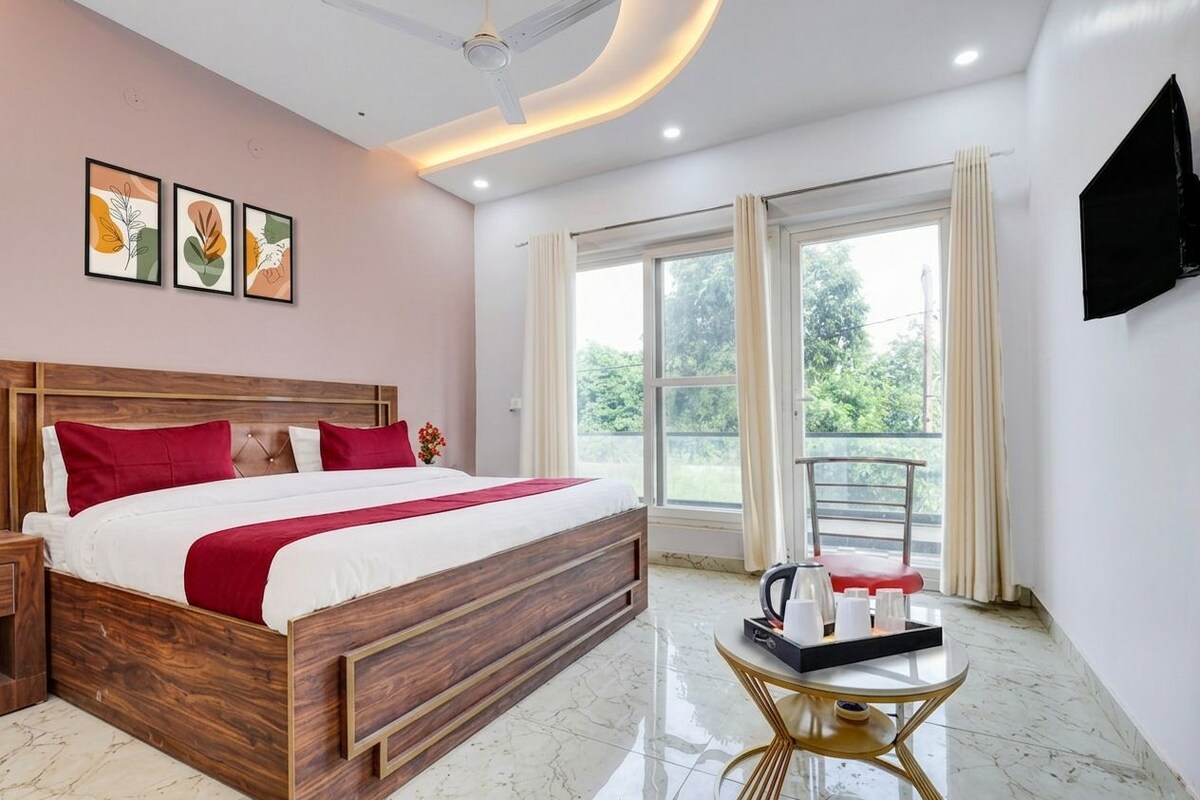 Quarto de hotel com cama de madeira, uma mesa redonda pequena com um tabuleiro, ventoinha no teto e uma janela grande com cortinas.
