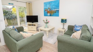 Uma sala de estar com sofás verdes, uma televisão de tela plana, um quadro de barco e um abajur.