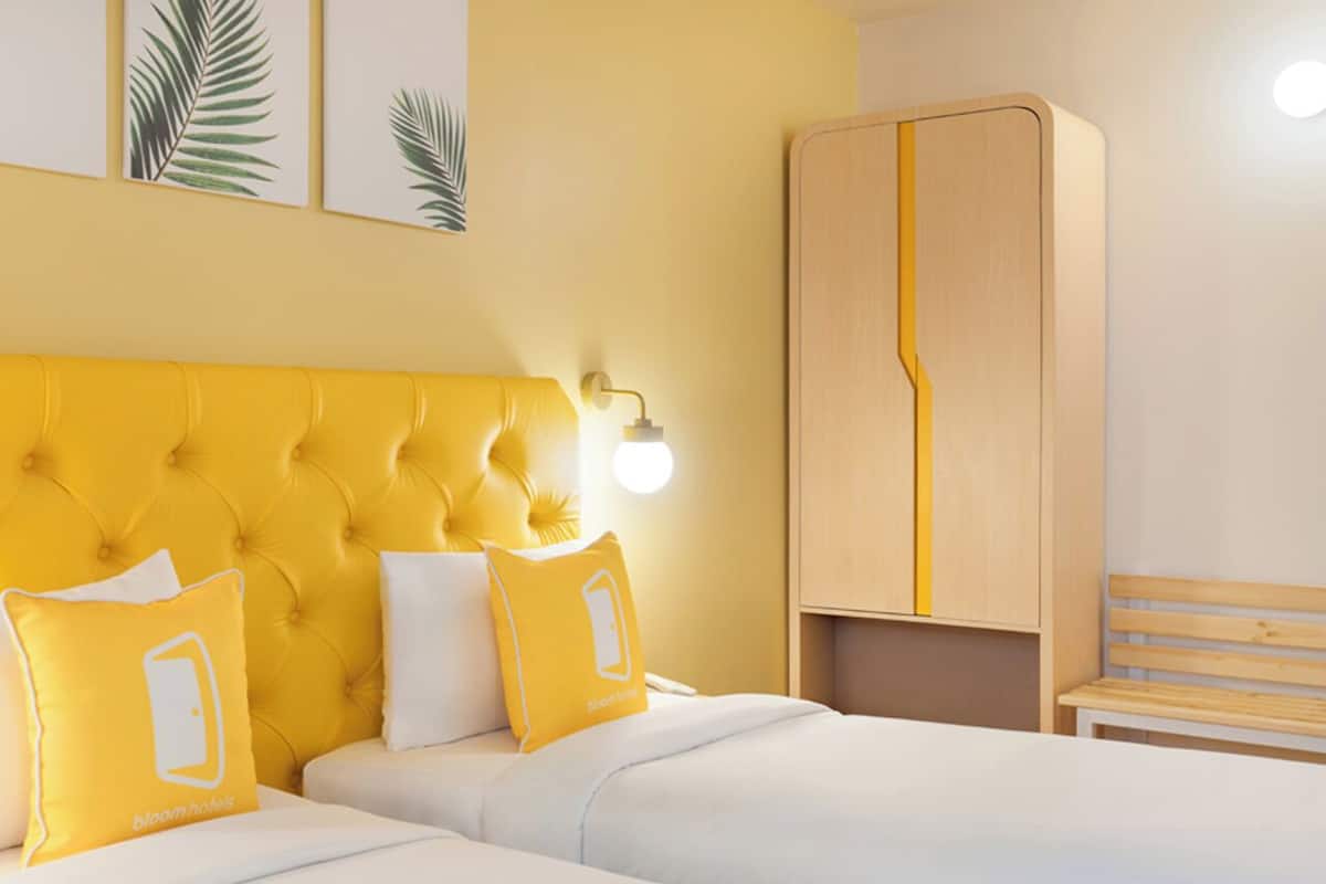 Quarto de hotel com cabeceira amarela acolchoada, duas camas com roupa de cama branca, almofadas amarelas e um guarda-roupa de madeira com detalhes amarelos.