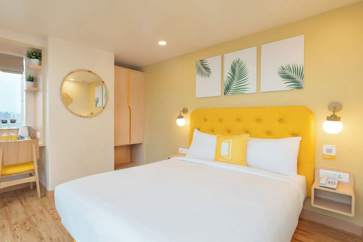 Quarto de hotel com uma cama grande, uma escrivaninha com cadeira, um espelho redondo e obras de arte na parede.