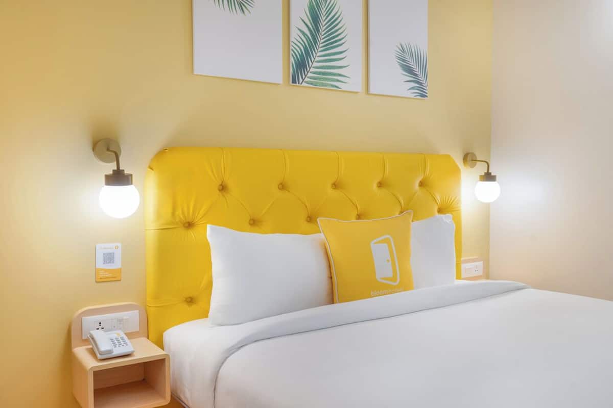 Quarto de hotel com cabeceira amarela com captonete, roupa de cama branca e mesa de cabeceira com telefone e abajur.