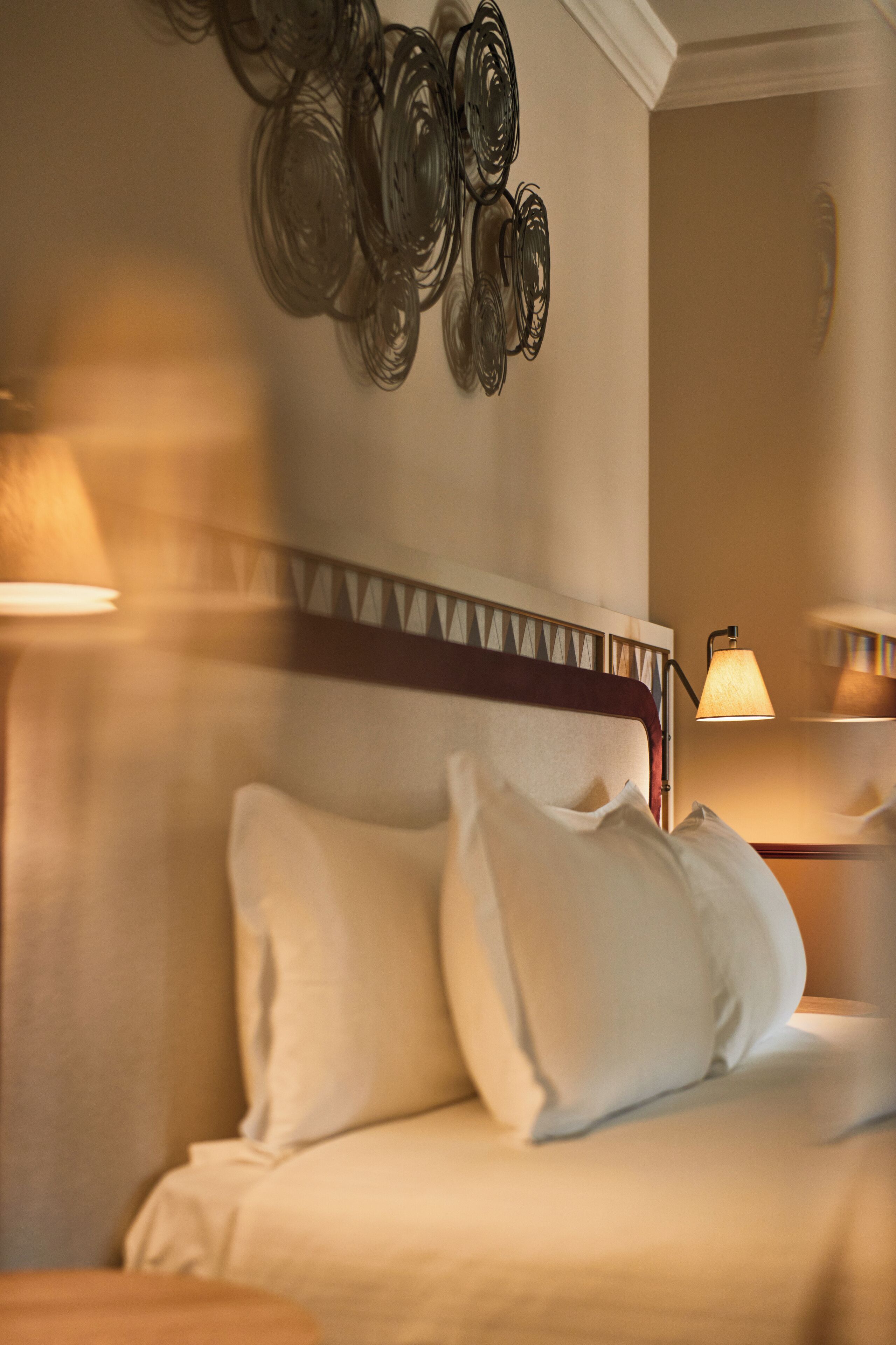 Quarto de hotel com uma cama, duas luminárias de cabeceira e um objeto decorativo na parede acima da cama.