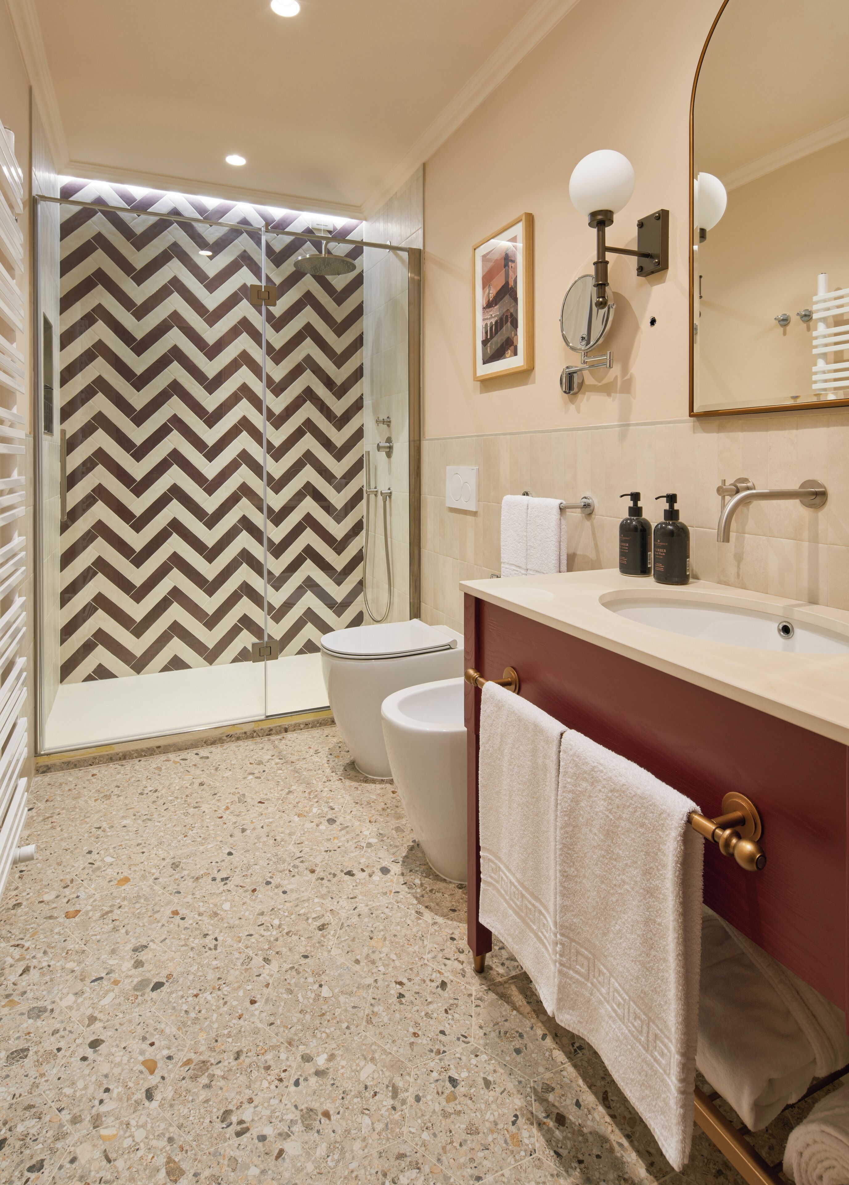 Casa de banho com chuveiro, sanita e lavatório, apresentando um padrão de mosaico herringbone nas paredes e soalho com mosaico salpicado.