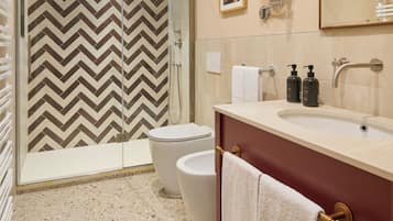 Casa de banho com chuveiro, sanita e lavatório, apresentando um padrão de mosaico herringbone nas paredes e soalho com mosaico salpicado.