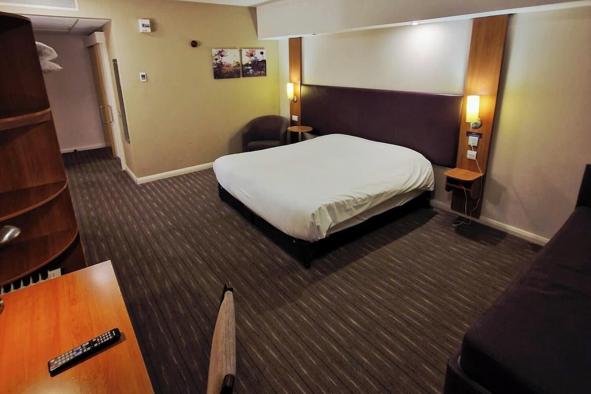 Quarto de hotel com cama, escrivaninha, cadeira, mesa pequena, mesa de cabeceira e luminária na parede.
