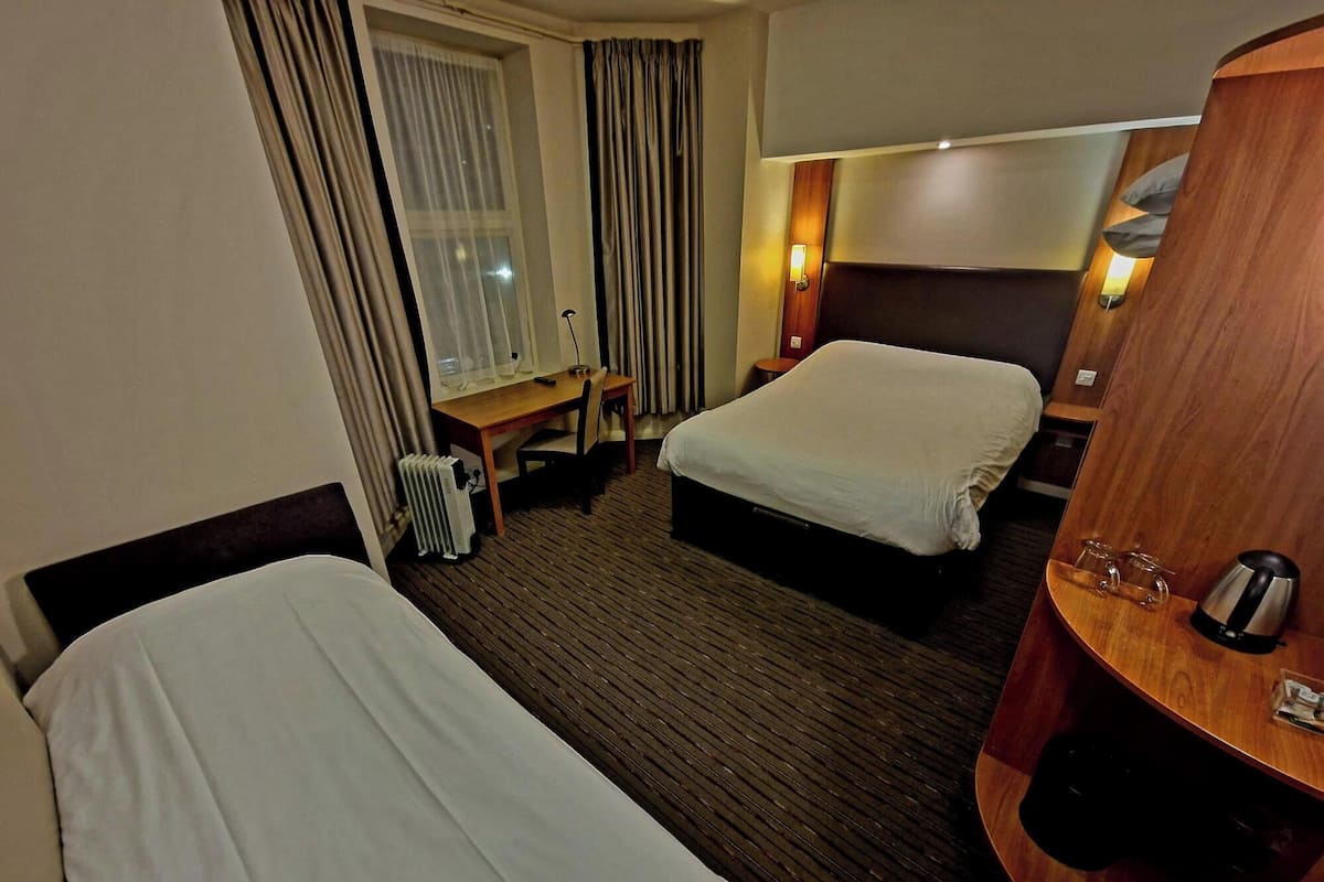 Quarto de hotel com duas camas, uma escrivaninha, uma cadeira, um radiador e uma mesa de cabeceira com luminária.