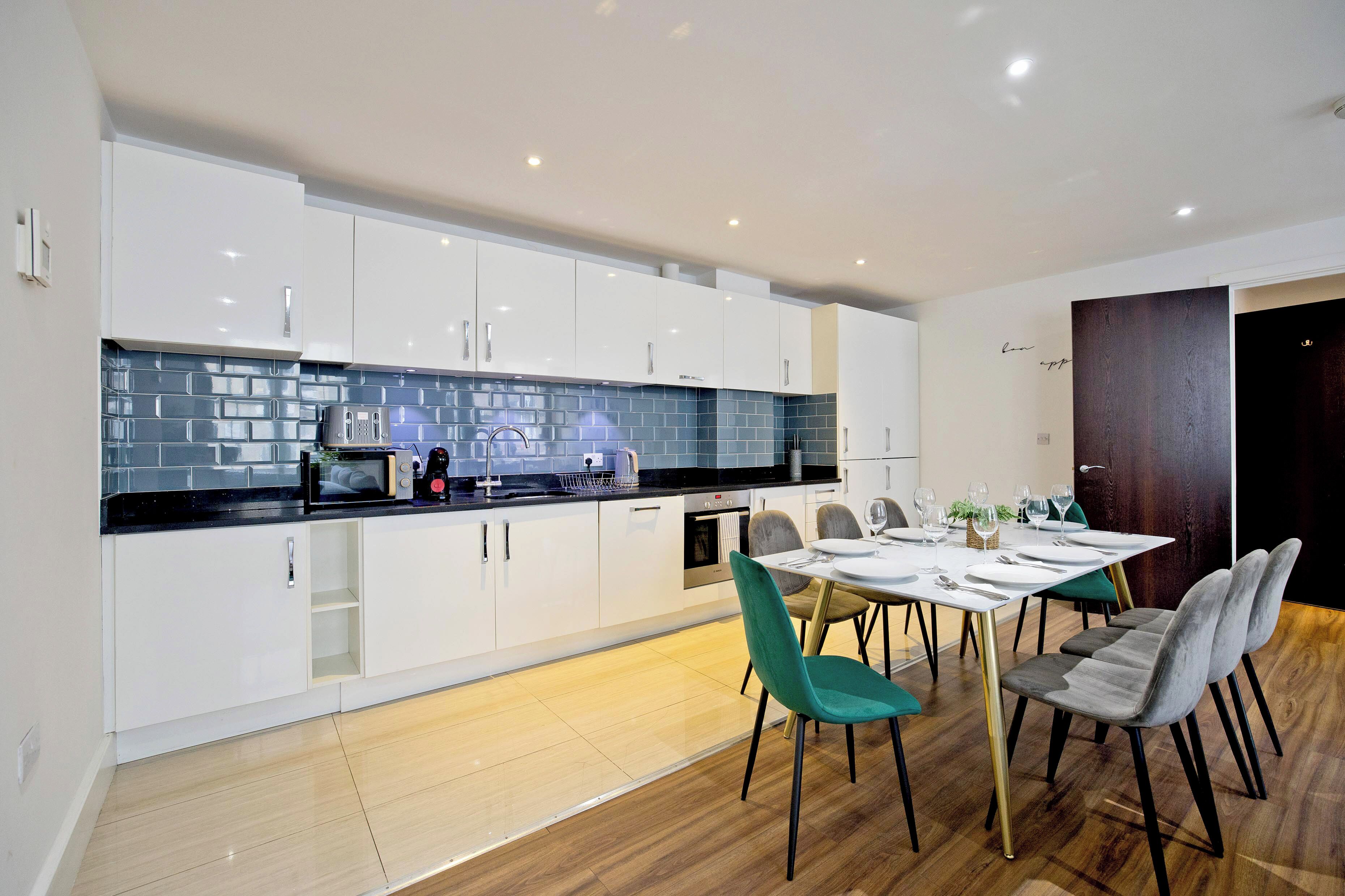 Cozinha moderna com armários brancos, bancada preta e painel de parede azul. Há uma mesa de jantar com cadeiras e piso de madeira.