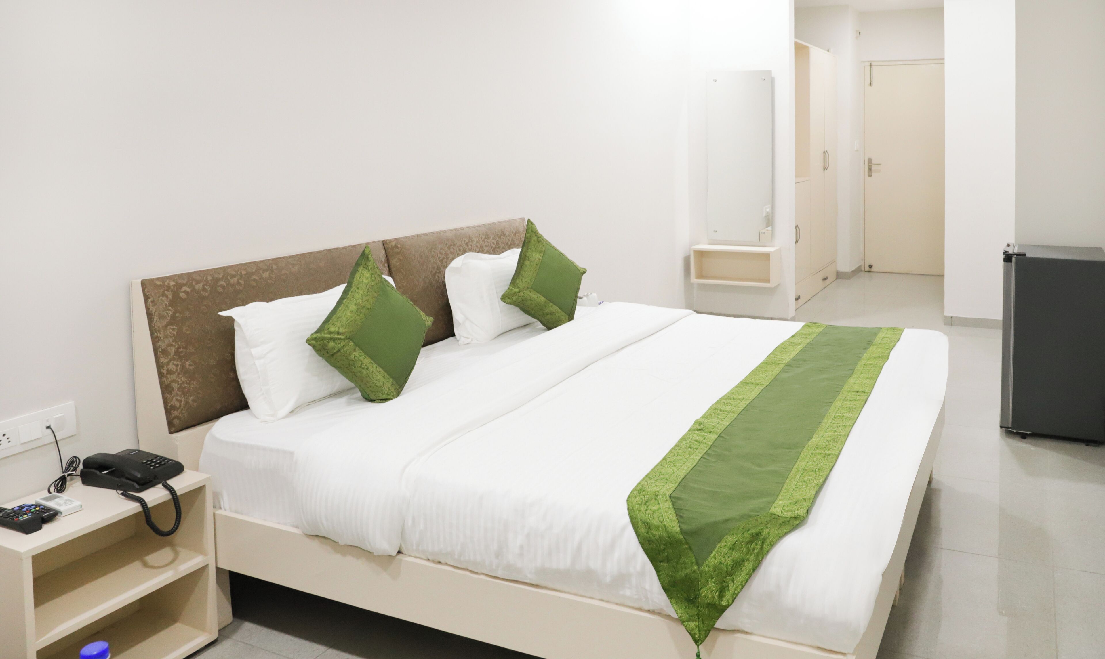 Uma cama bem arrumada com roupa de cama branca e detalhes verdes, um telefone ao lado da cama e uma pequena prateleira com um telefone e carregador.