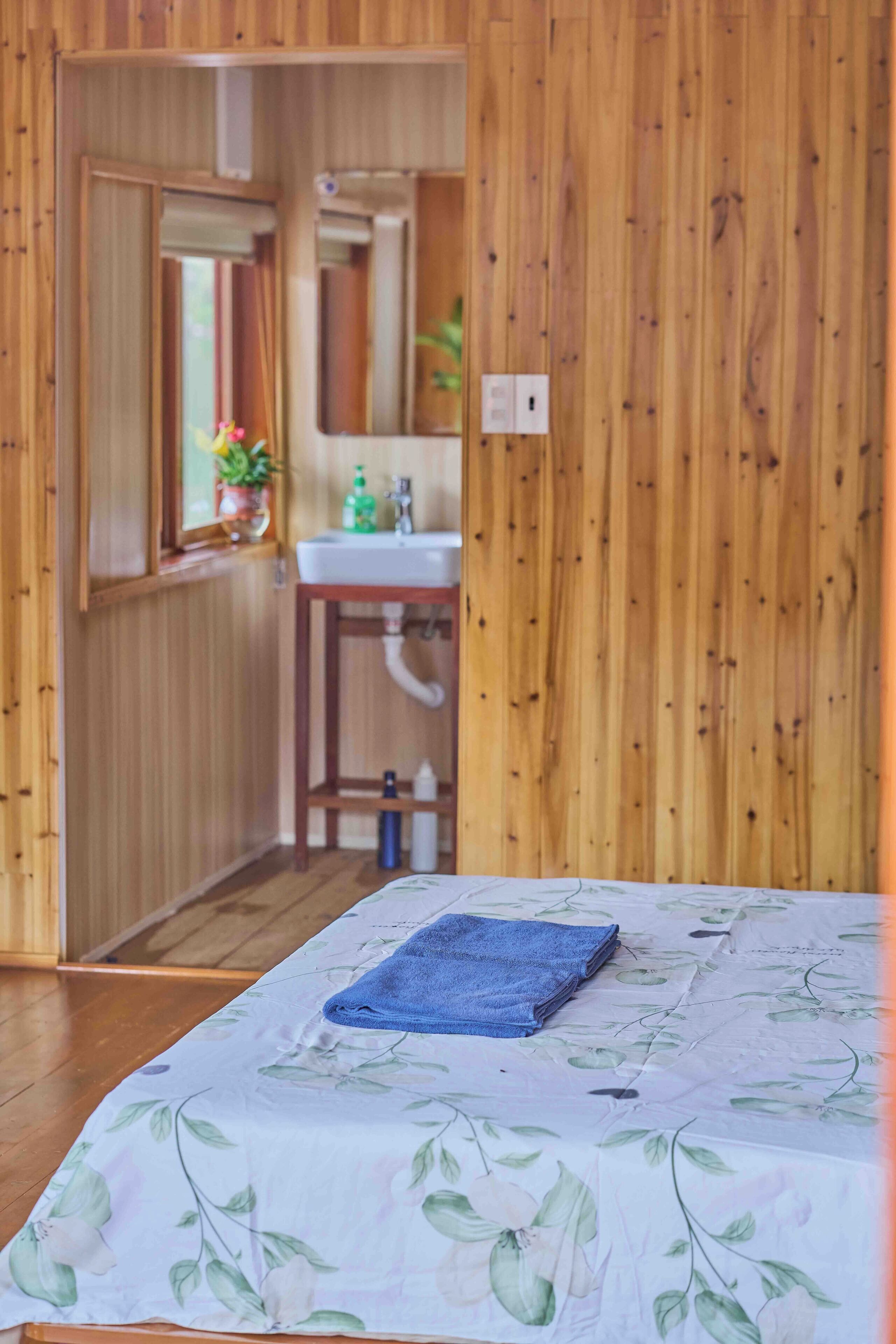 Quarto com painéis de madeira, cama com colcha floral, mesa pequena com toalha azul e banheiro visível através de uma porta aberta.