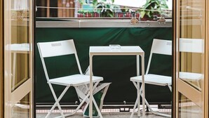 Visão através de uma porta de vidro de um quarto com duas cadeiras brancas e uma pequena mesa, uma cortina verde e um edifício de tijolos ao fundo.