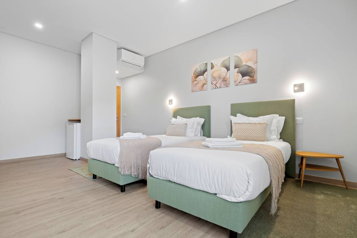 Quarto de hotel moderno com duas camas, uma mesinha e ar-condicionado fixado na parede.