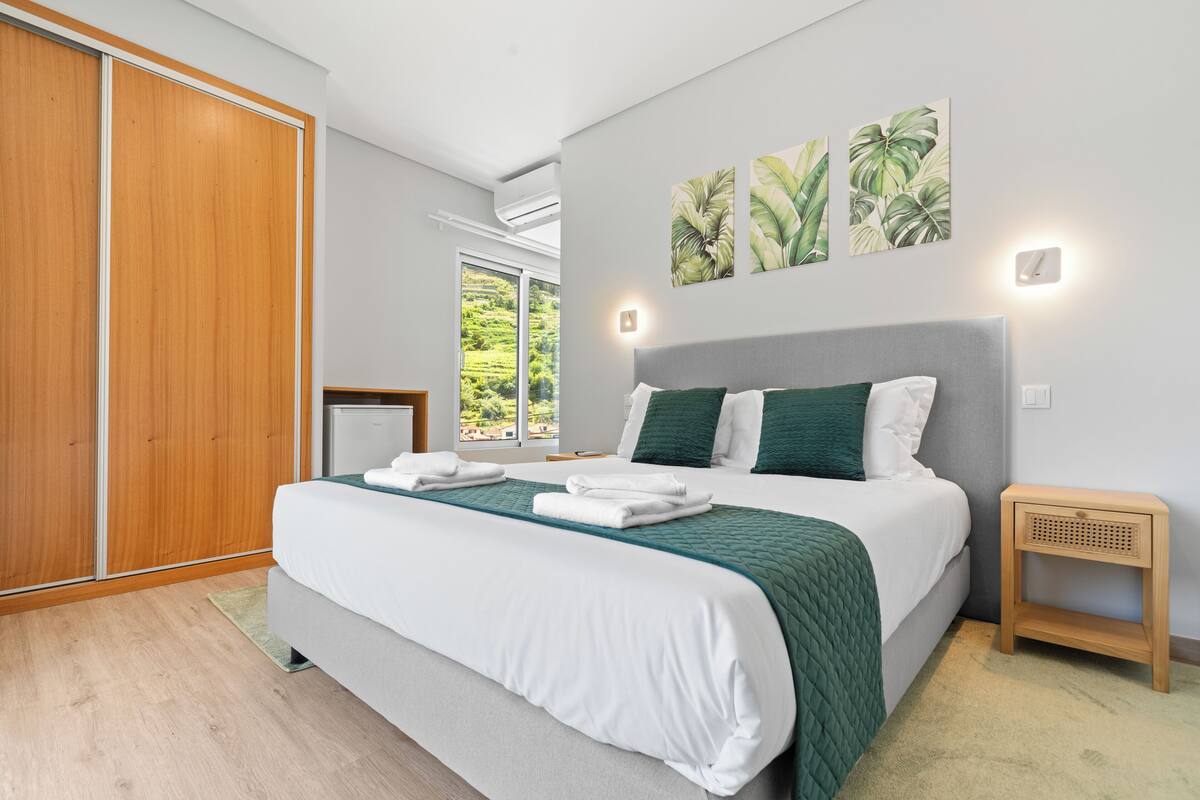 Quarto moderno com cama grande, guarda-roupa e vista para o verde pela janela.