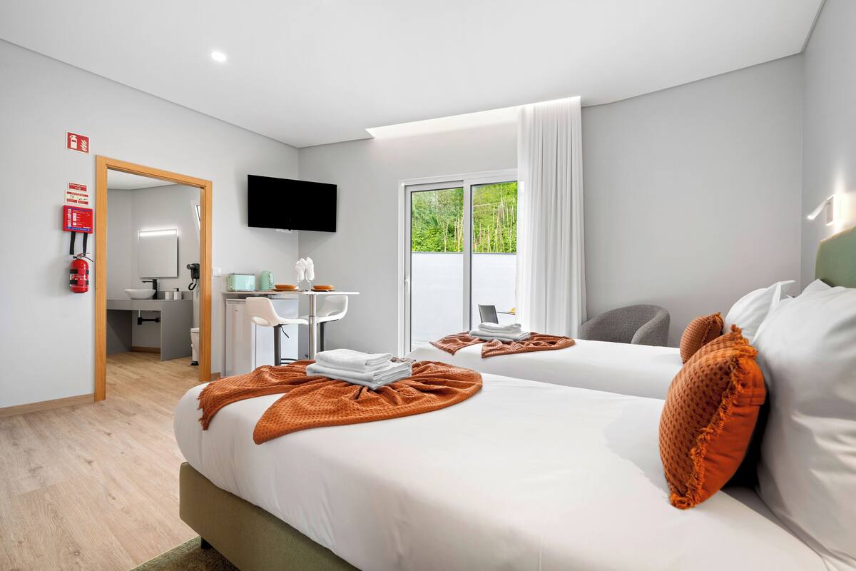 Quarto de hotel com duas camas, uma escrivaninha, televisão e janela com vista para o verde.