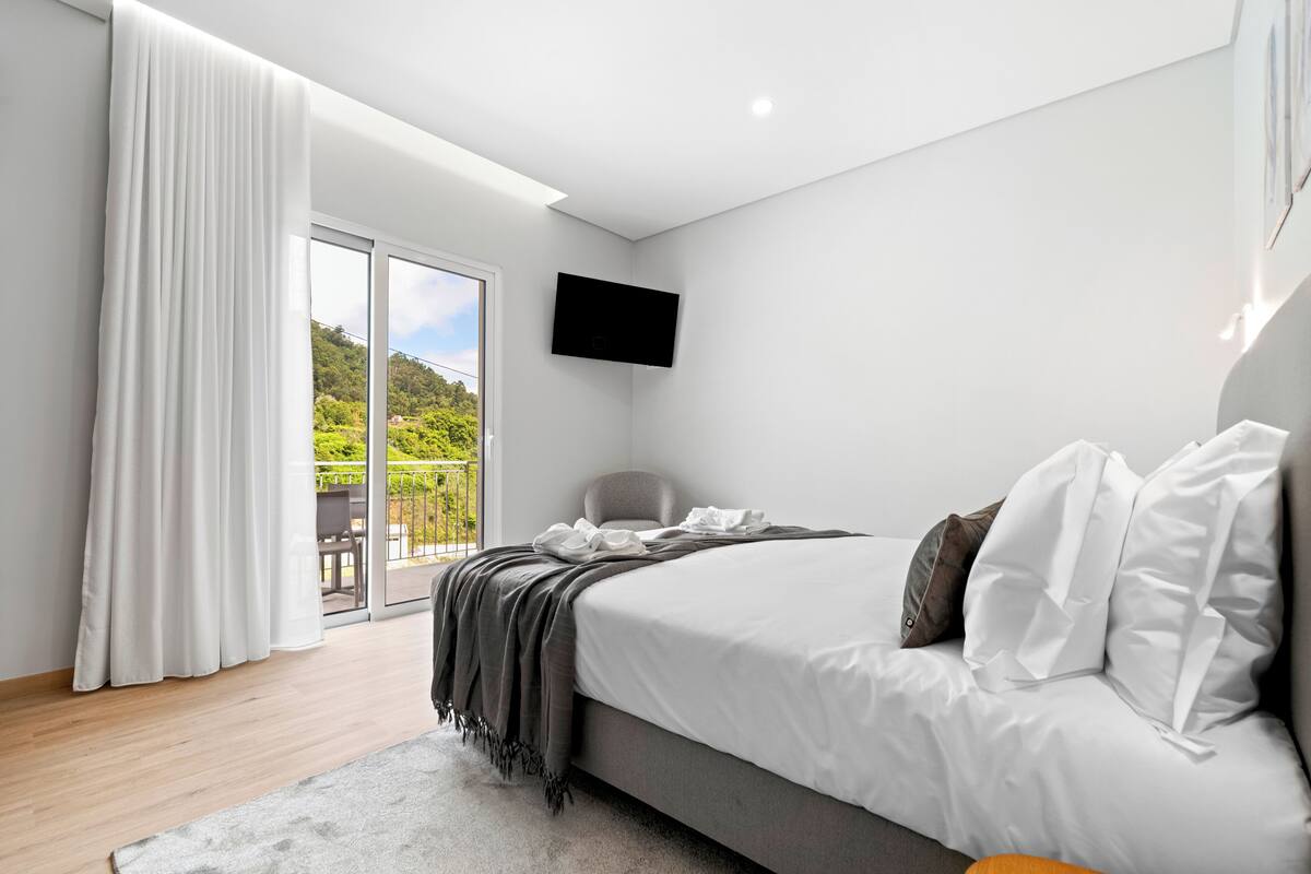Quarto moderno com uma cama grande, televisão de tela plana e varanda com vista para o verde.