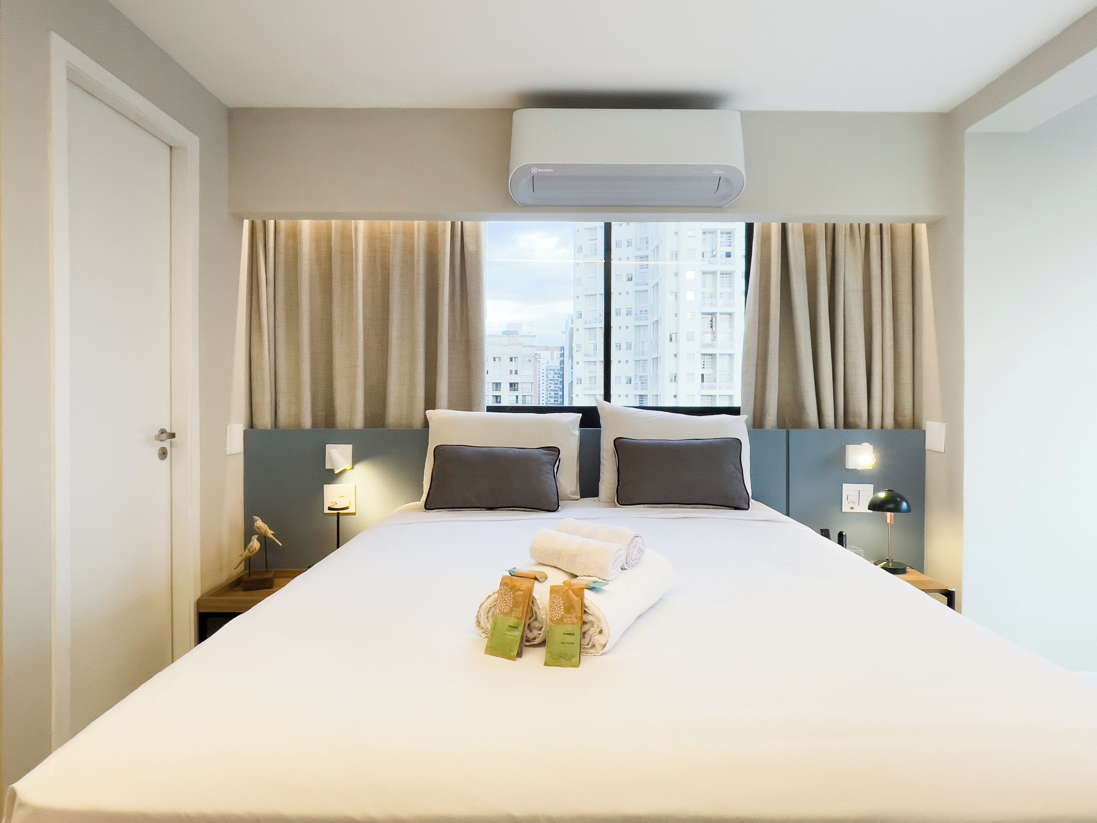 Quarto de hotel com uma cama grande, duas almofadas, uma mesinha de cabeceira com abajur e uma janela com cortinas.