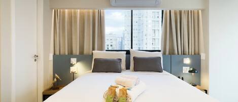 Quarto de hotel com uma cama grande, duas almofadas, uma mesinha de cabeceira com abajur e uma janela com cortinas.