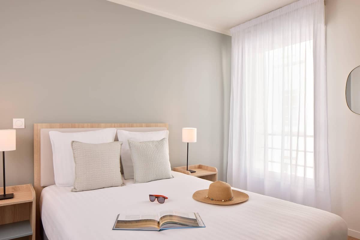 Cama bem arrumada com um livro, óculos de sol e um chapéu de palha. Há duas luminárias de cabeceira e uma mesa de cabeceira com um abajur.