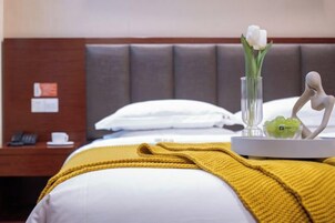 Quarto de hotel com uma cama, mesa de cabeceira com telefone, um vaso de flores e um objeto decorativo na mesa.