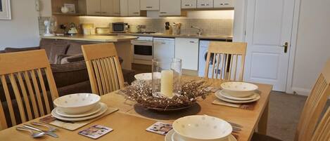 Uma área de refeições com uma mesa de madeira posta para quatro pessoas, cercada por cadeiras. Um centro de mesa decorativo com uma vela está no meio.