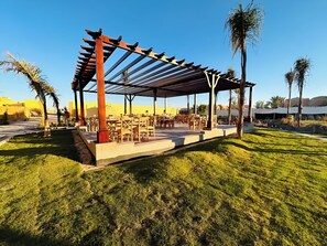 Área externa com mesas e cadeiras sob uma pérgola, cercada por grama e palmeiras.