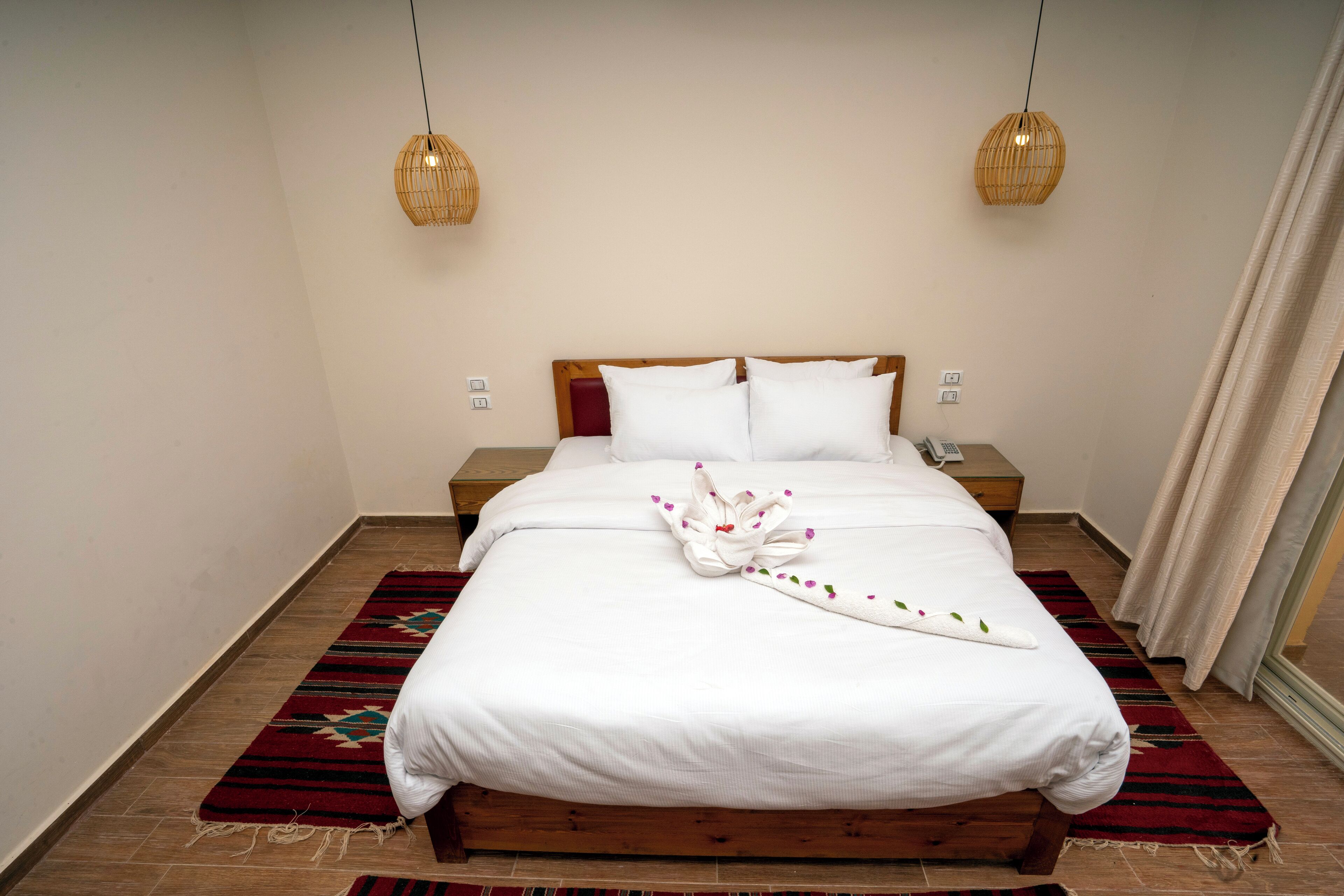 Cama bem arrumada com lençóis brancos e toalha disposta em padrão floral, cabeceira de madeira, duas mesas de cabeceira com luminárias e um tapete listrado em vermelho e preto no chão.
