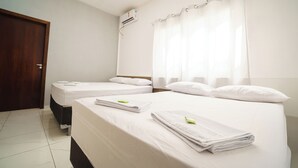 Quarto de hotel com duas camas, roupa de cama branca, porta de madeira, unidade de ar condicionado e janela com cortinas brancas.