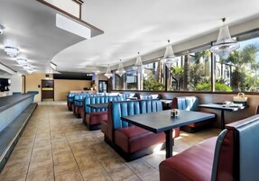Um restaurante com cabines e mesas, grandes janelas e vista para palmeiras.