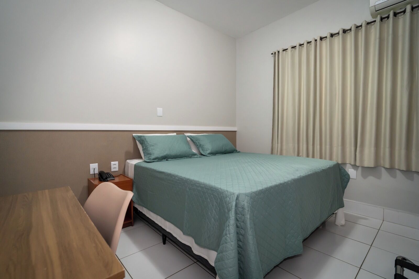 Quarto de hotel com cama, mesa de cabeceira, cadeira, janela com cortinas e radiador.