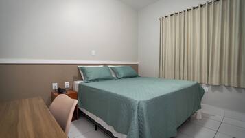 Quarto de hotel com cama, mesa de cabeceira, cadeira, janela com cortinas e radiador.