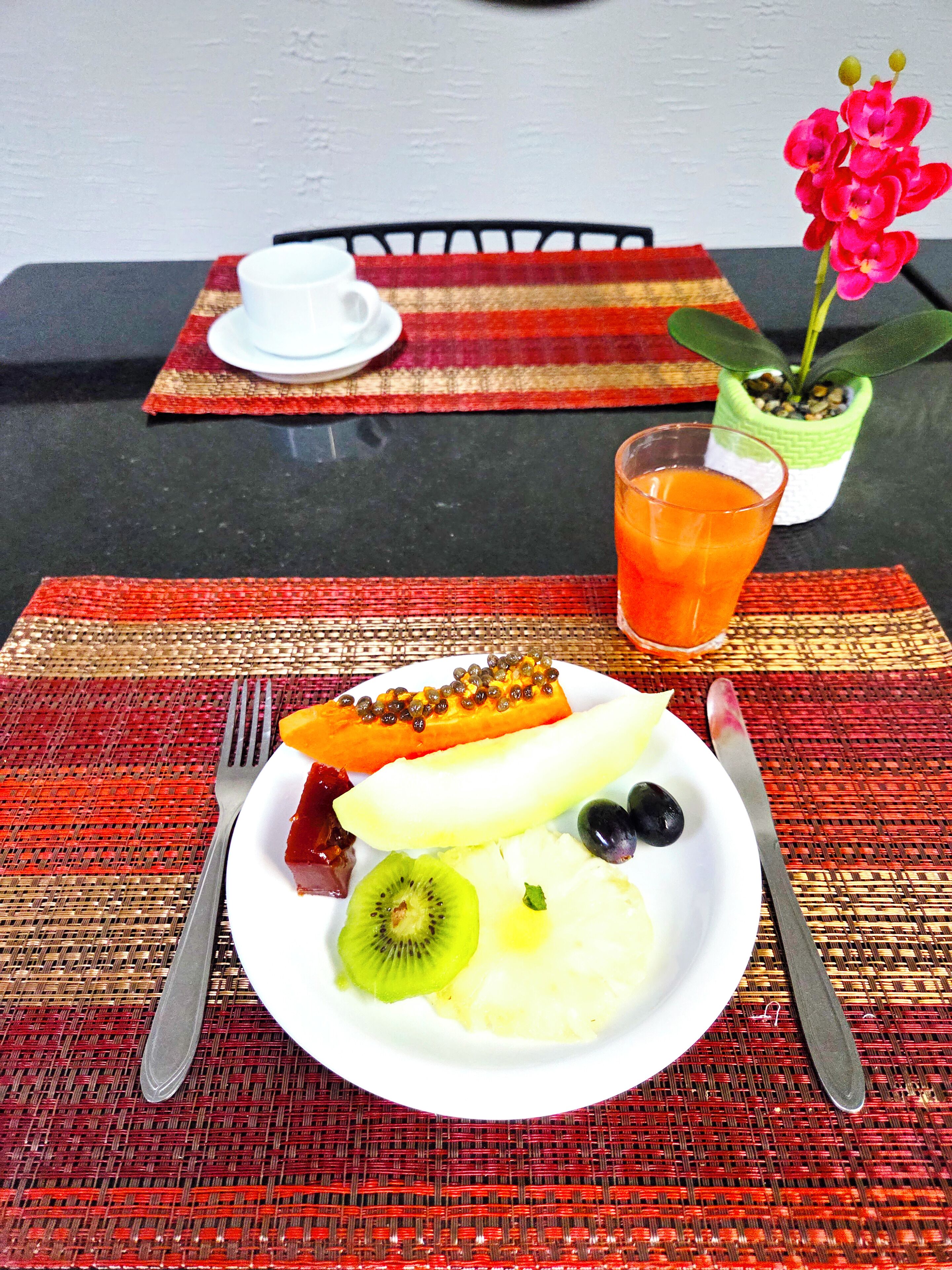 Um cenário de café da manhã com uma travessa de frutas, um copo de suco de laranja e um pequeno vaso de planta.