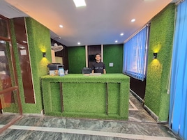 Um balcão de recepção com uma superfície de grama sintética verde, um computador e um ventilador. Duas luminárias fixadas na parede em ambos os lados do balcão.