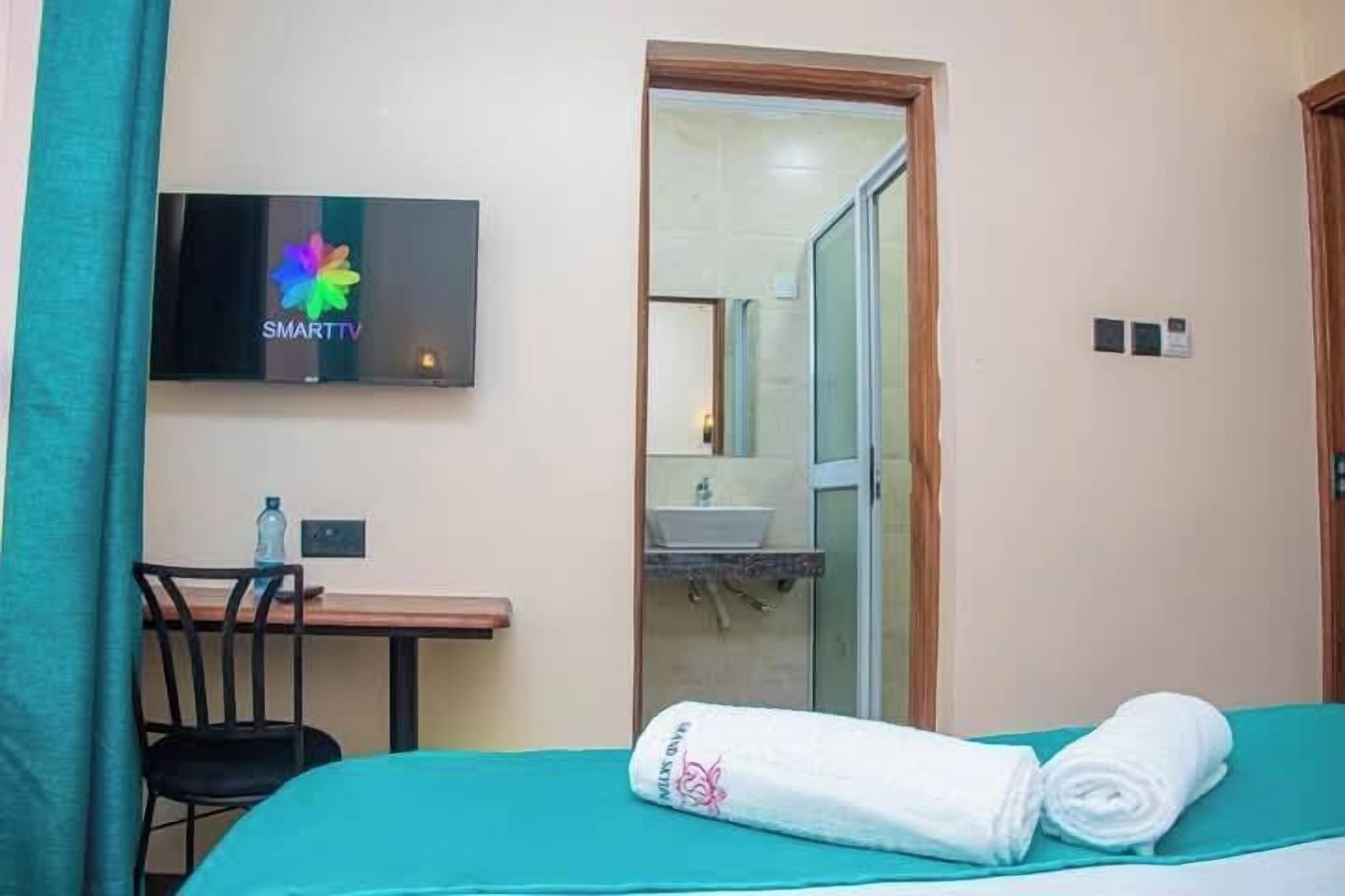 Quarto de hotel com cama, escrivaninha, cadeira, TV fixada na parede e banheiro visível por uma porta aberta.