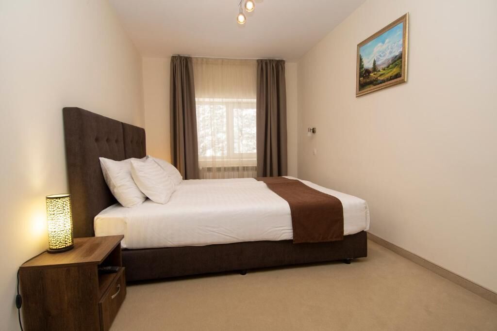 Quarto com cama, mesa de cabeceira com abajur, janela com cortinas e um quadro na parede.