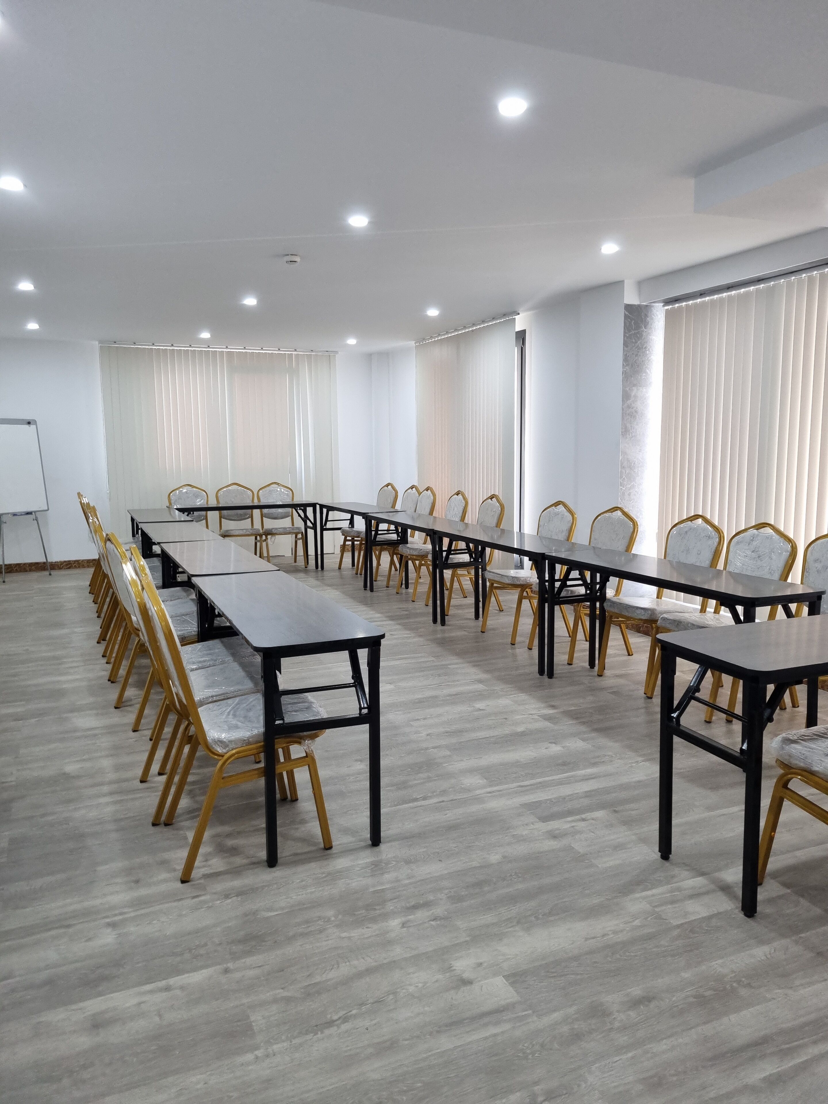 Uma sala de conferências com mesas compridas e cadeiras, paredes brancas e janelas grandes com persianas.