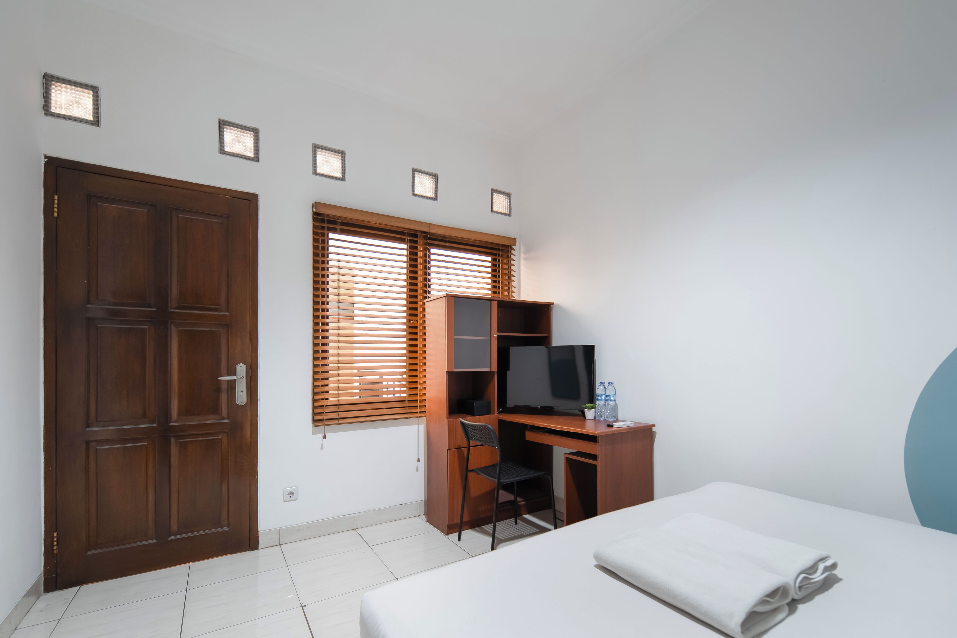 Quarto de hotel com porta de madeira, escrivaninha com televisão, cama e janela com persianas.