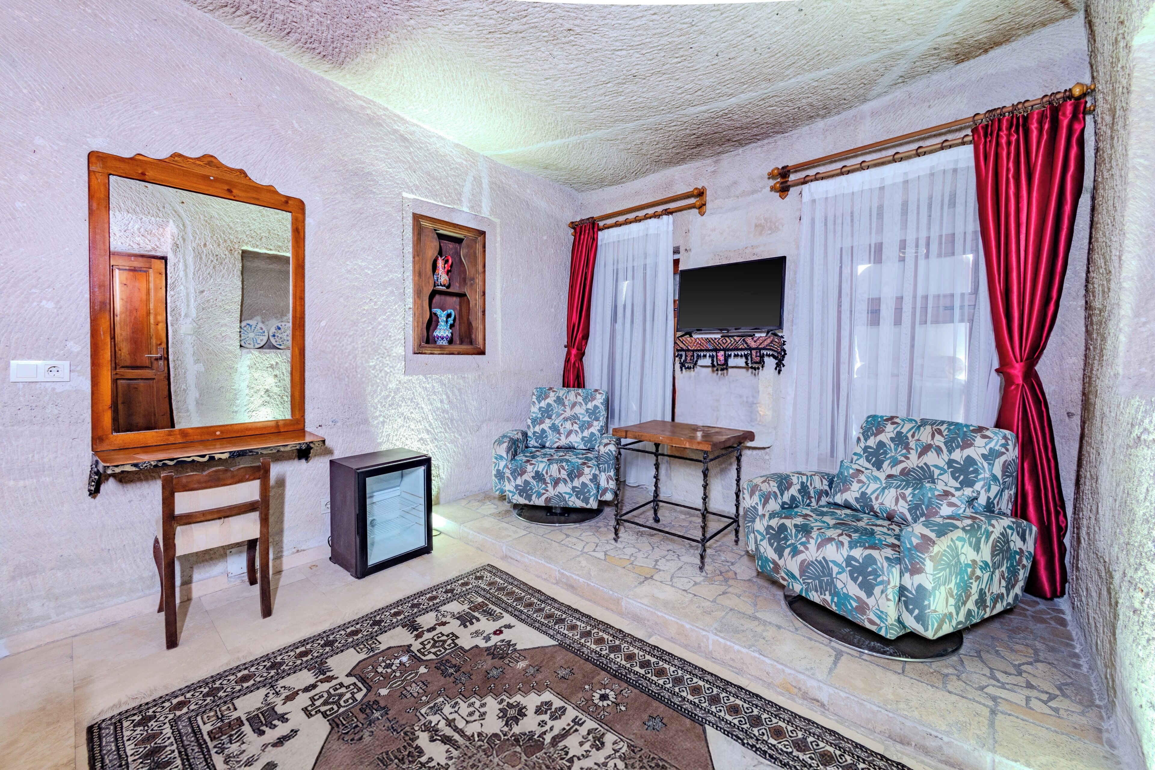 Uma sala de estar com uma cadeira de madeira, um sofá, uma mesinha, uma televisão e um tapete estampado.