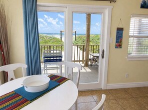 Quarto com mesa, cadeiras e vista para a praia, além de porta que dá para o exterior.