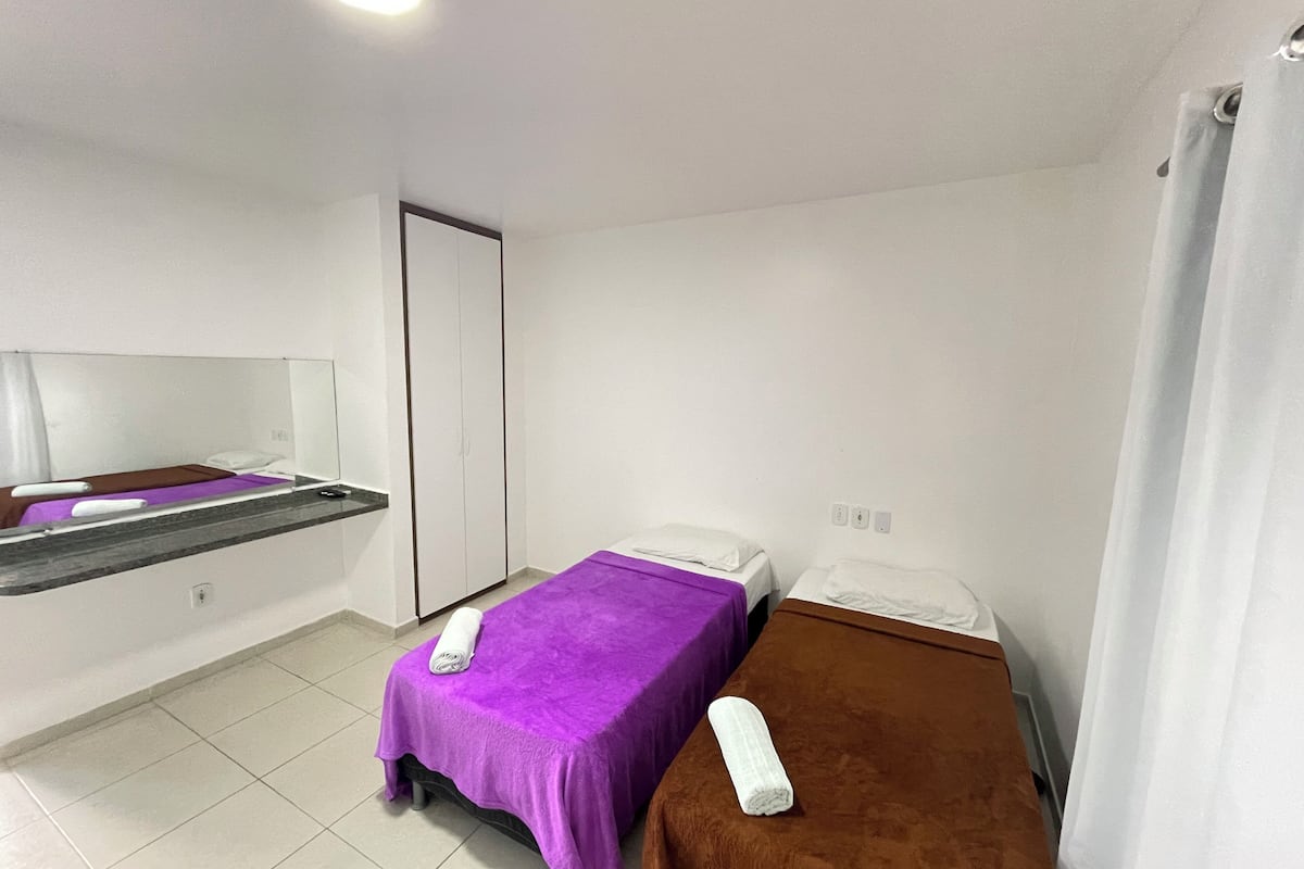 Quarto com duas camas, uma com colcha roxa e outra com colcha marrom. Há um espelho na parede, uma cortina branca e uma porta branca.