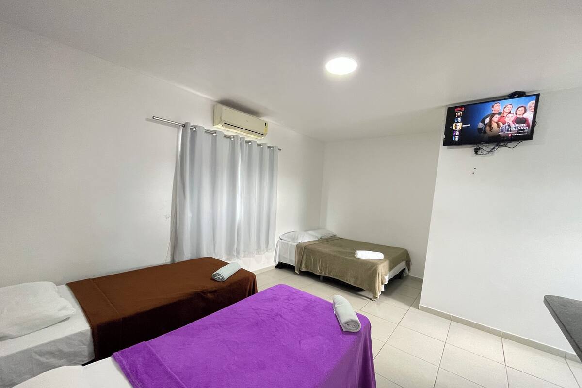 Quarto de hotel com duas camas, TV fixada na parede, janela com cortinas e ventoinha no teto.