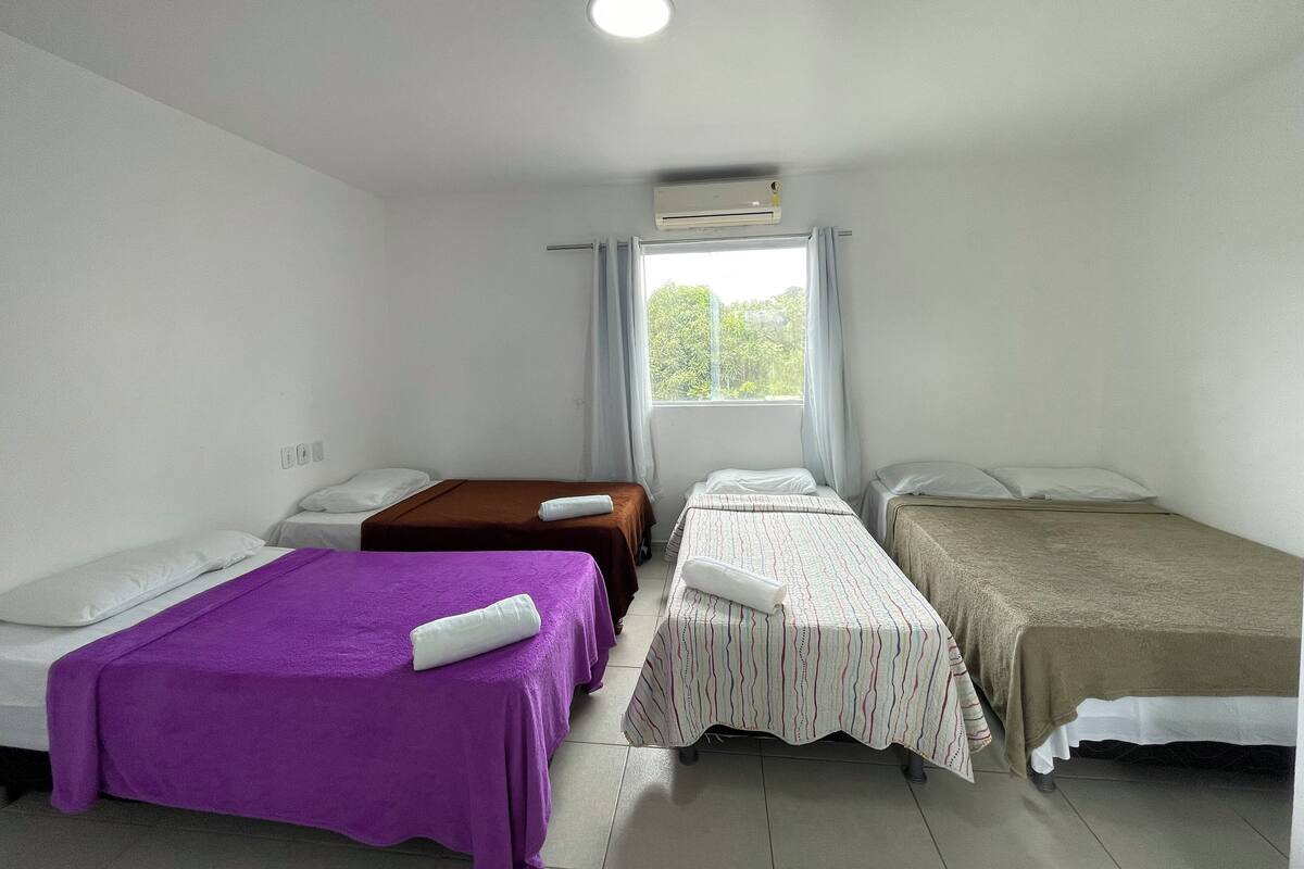 Quarto com três camas de solteiro, cada uma com colcha de cor diferente. Há uma janela com cortinas, um aparelho de ar condicionado e uma luminária no teto.