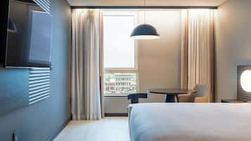 Quarto de hotel moderno com uma cama grande, uma escrivaninha, uma cadeira, uma televisão e uma janela com cortinas.