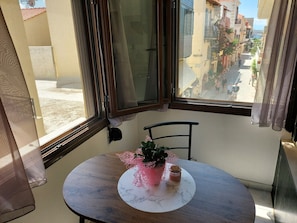 Quarto com mesa redonda, uma cadeira, janela com cortinas e vista para a rua com edifícios.
