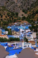 Um povoado em encosta densamente construído, com edifícios em tons variados de azul e branco, combinando arquitetura tradicional e moderna.