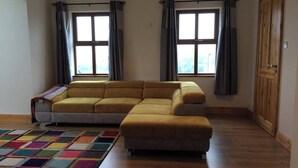 Uma sala de estar com sofá modular, duas janelas com cortinas, um tapete colorido e uma porta de madeira.