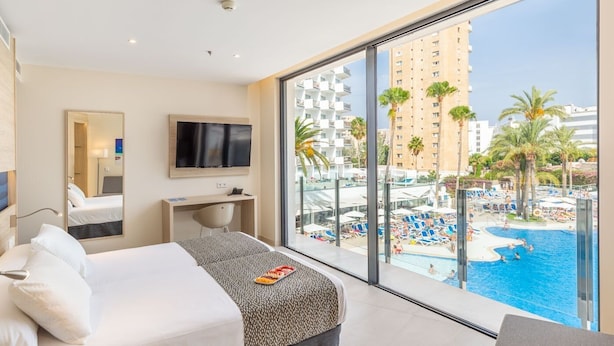 Quarto de hotel com uma janela grande, uma cama, uma escrivaninha e uma televisão.