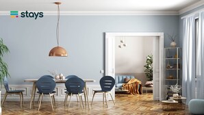 Uma área de jantar moderna com chão de madeira em padrão herringbone, um pendente de cobre e um sofá azul ao fundo.