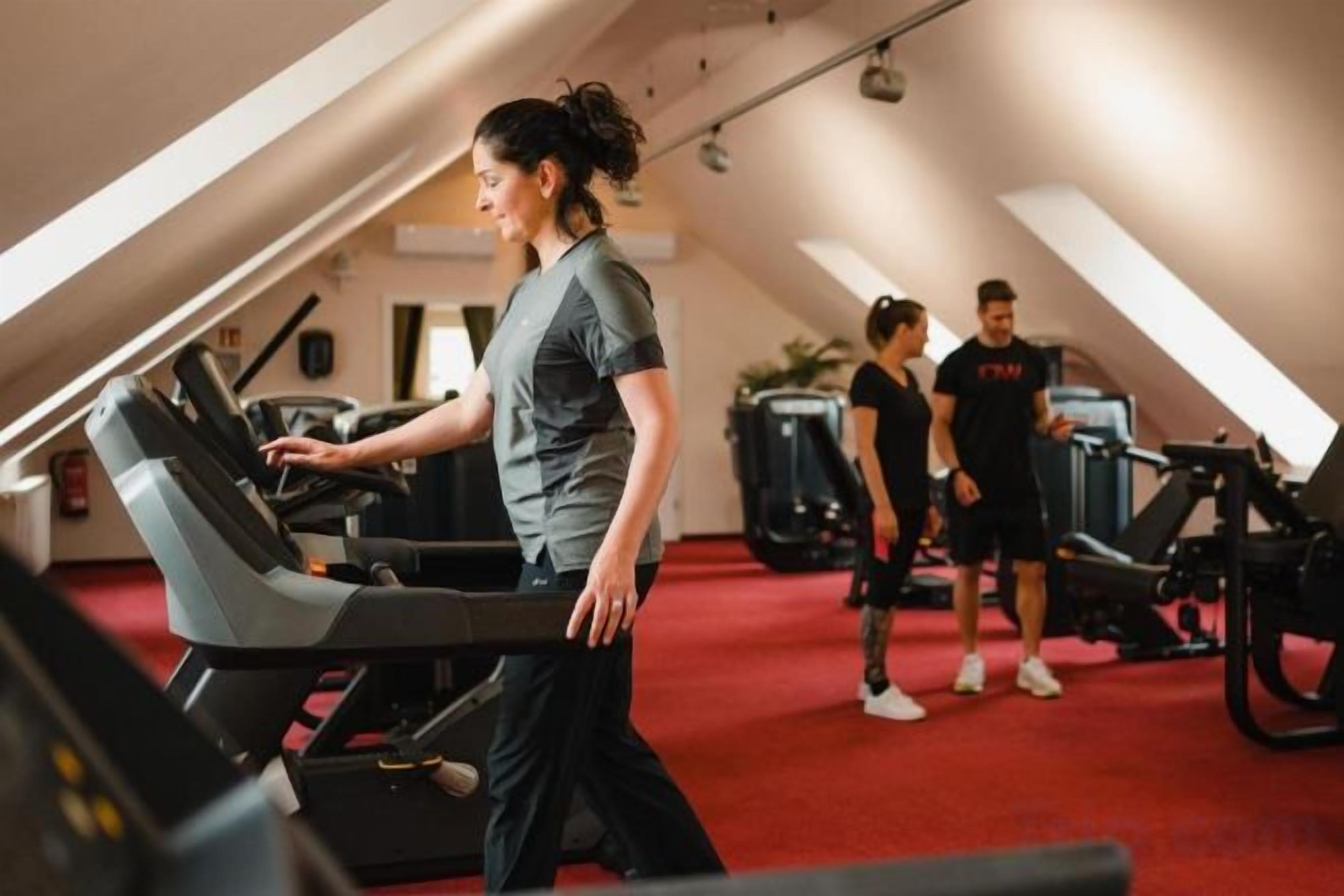 Uma academia com esteiras, equipamentos de exercício e piso coberto por carpete vermelho.